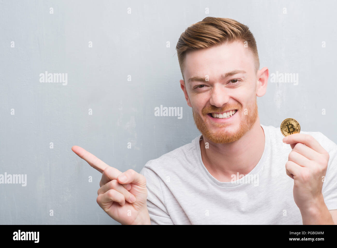Young redhead man over grey grunge wall showing bitcoin very happy ...