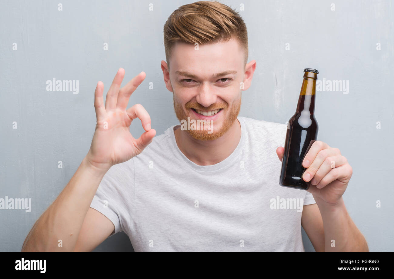 Young redhead man over grey grunge wall drinking beer bottle doing ok ...
