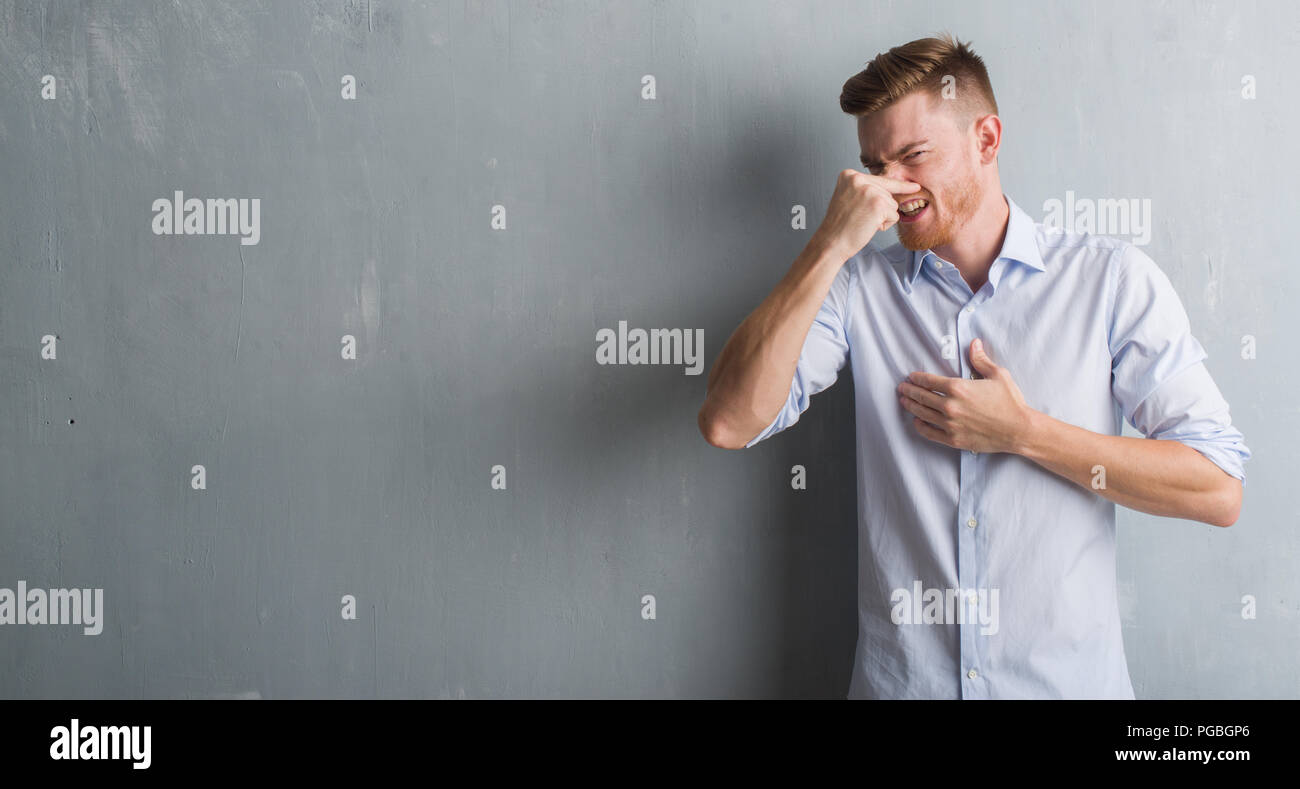 Young redhead business man over grey grunge wall smelling something ...