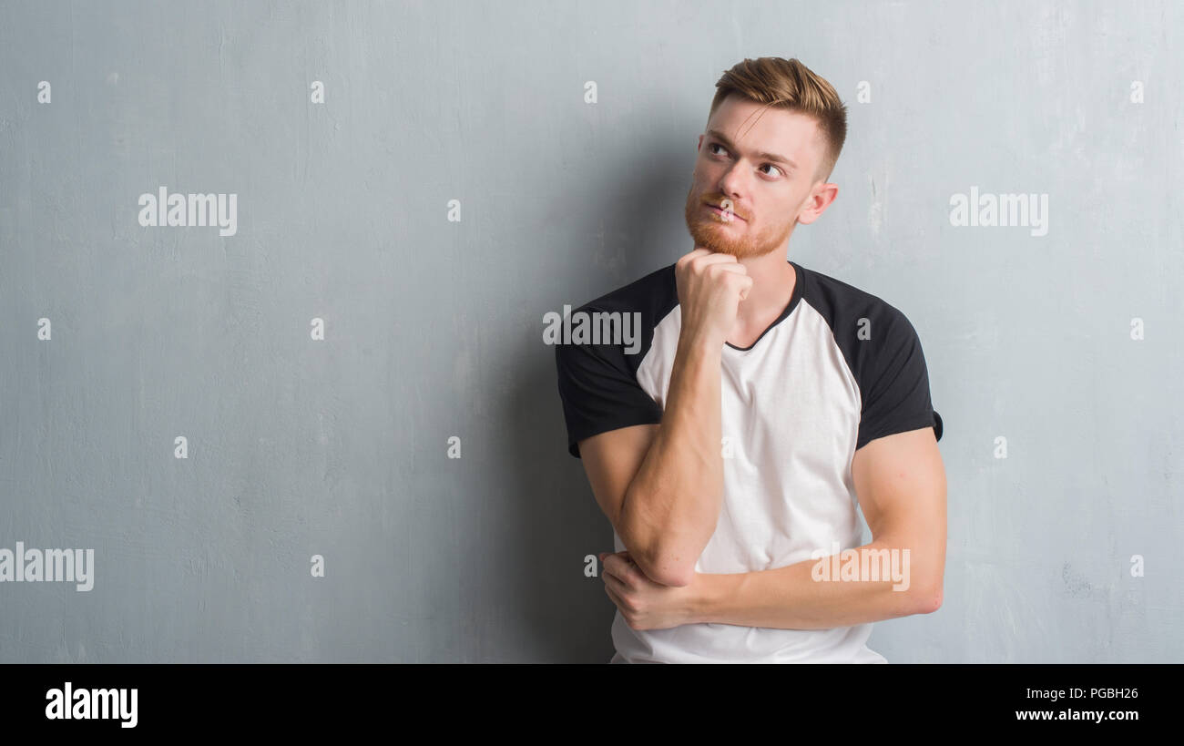 Young redhead man over grey grunge wall with hand on chin thinking ...