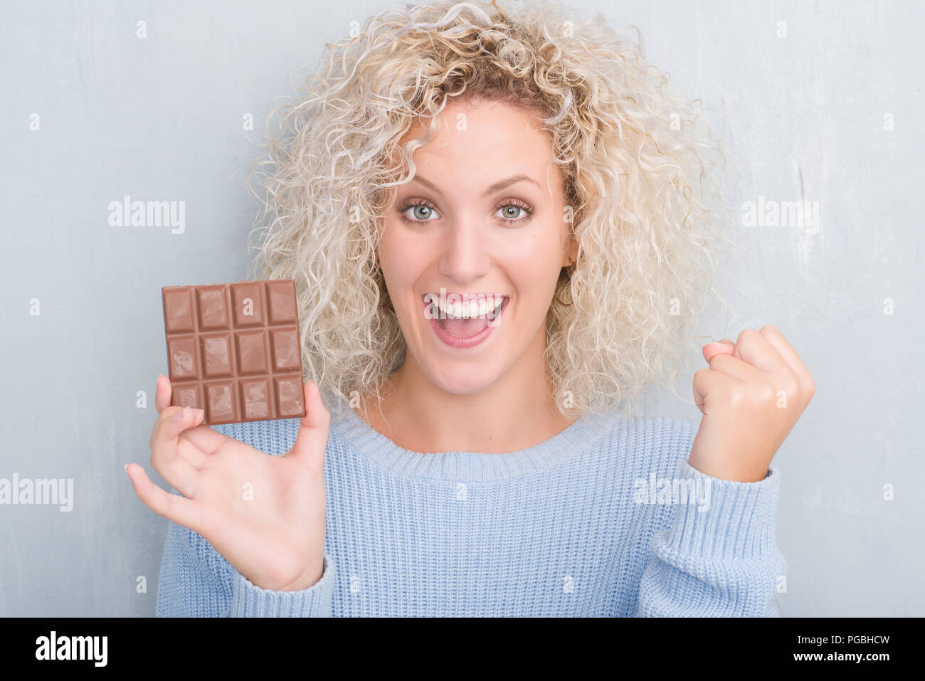 Young blonde woman over grunge grey wall eating chocolate bar screaming ...