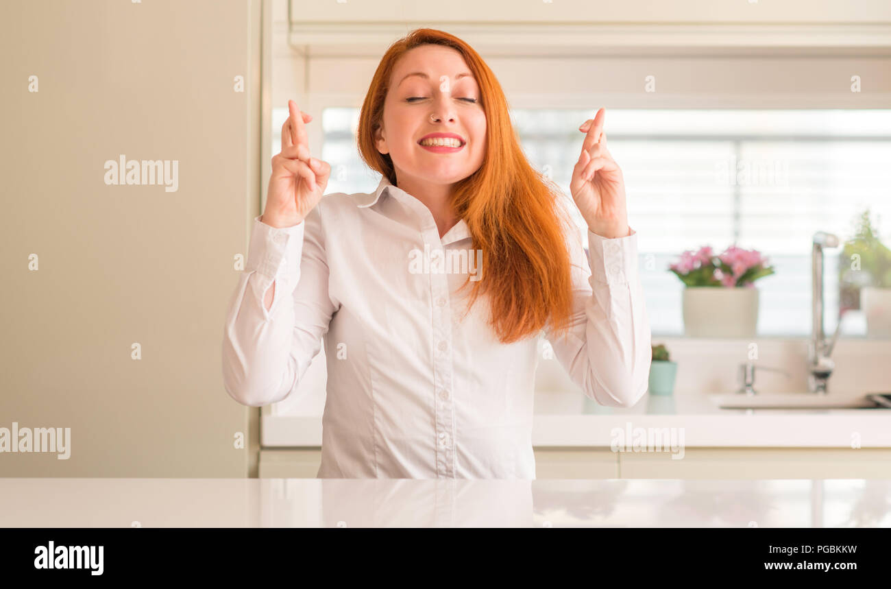 Redhead woman at kitchen smiling crossing fingers with hope and eyes ...