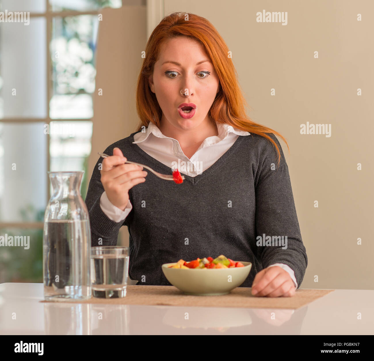 Redhead woman eating fruit bowl, kiwi and strawberry at home scared in ...