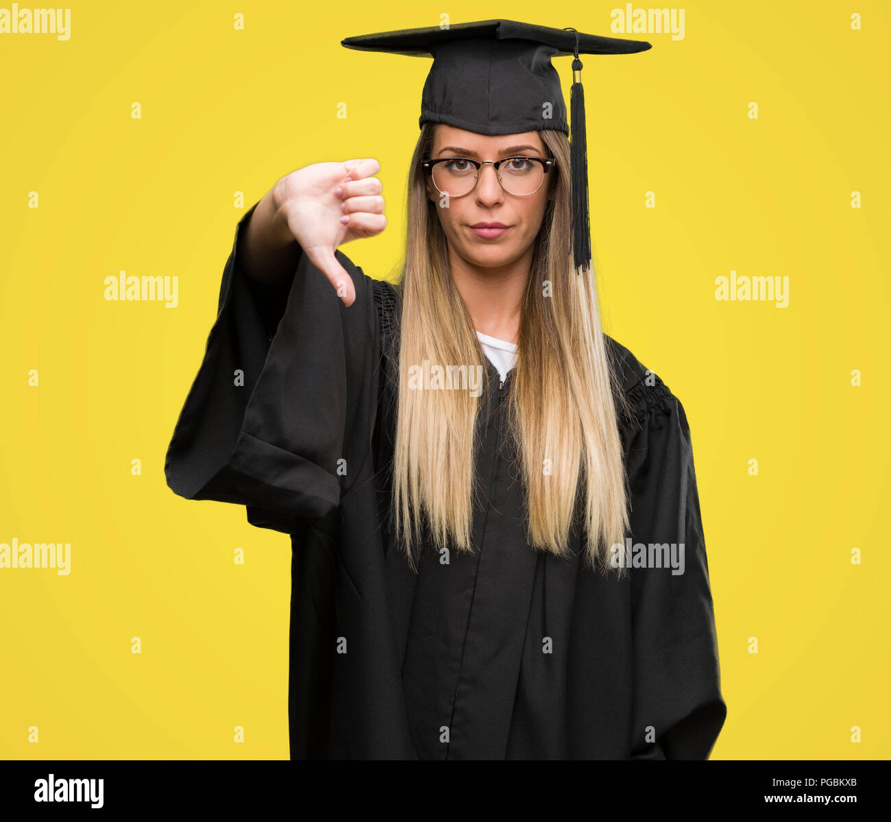 Beautiful young woman wearing glasses and graduation robe with angry ...