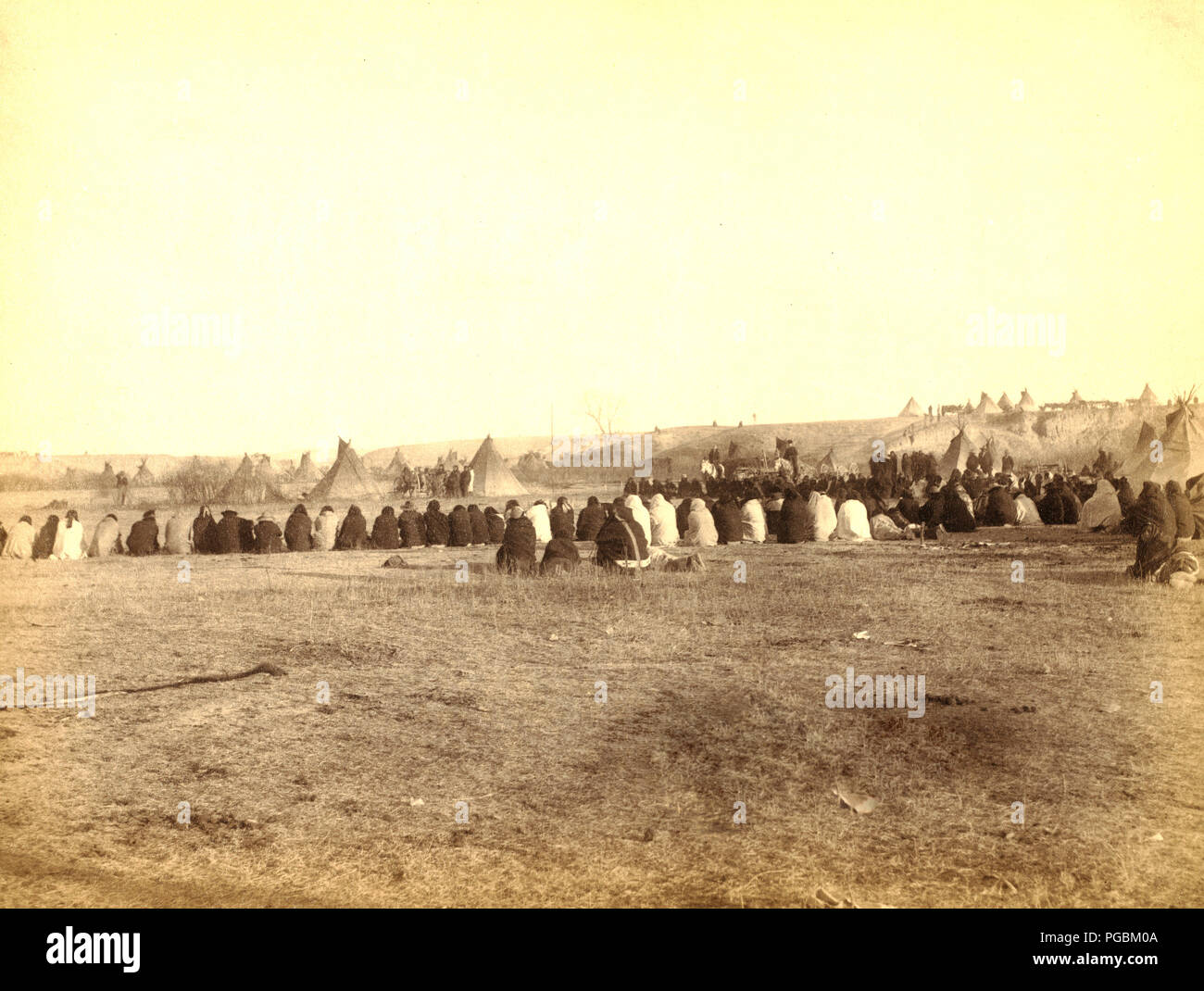 Rear view of a large semi-circle of Lakota men sitting on the ground ...