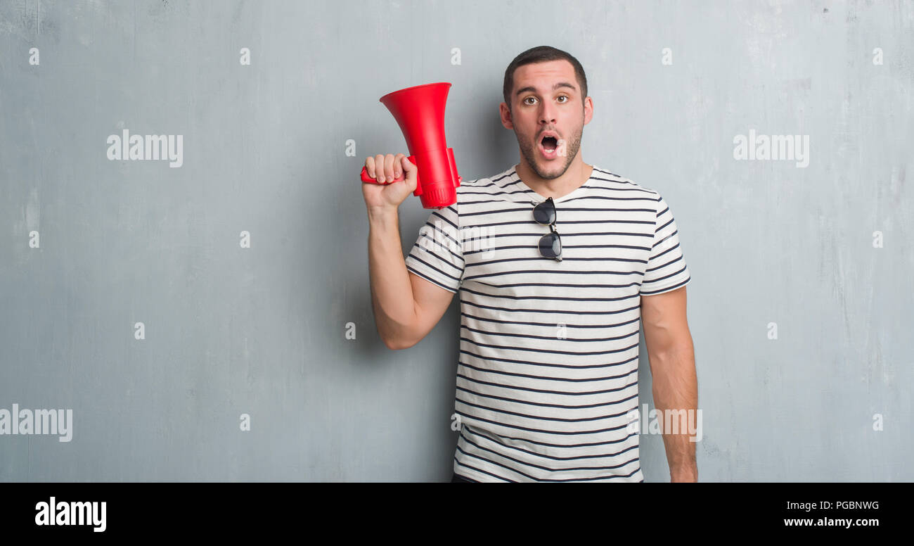 Young caucasian man over grey grunge wall shouting trough megaphone ...