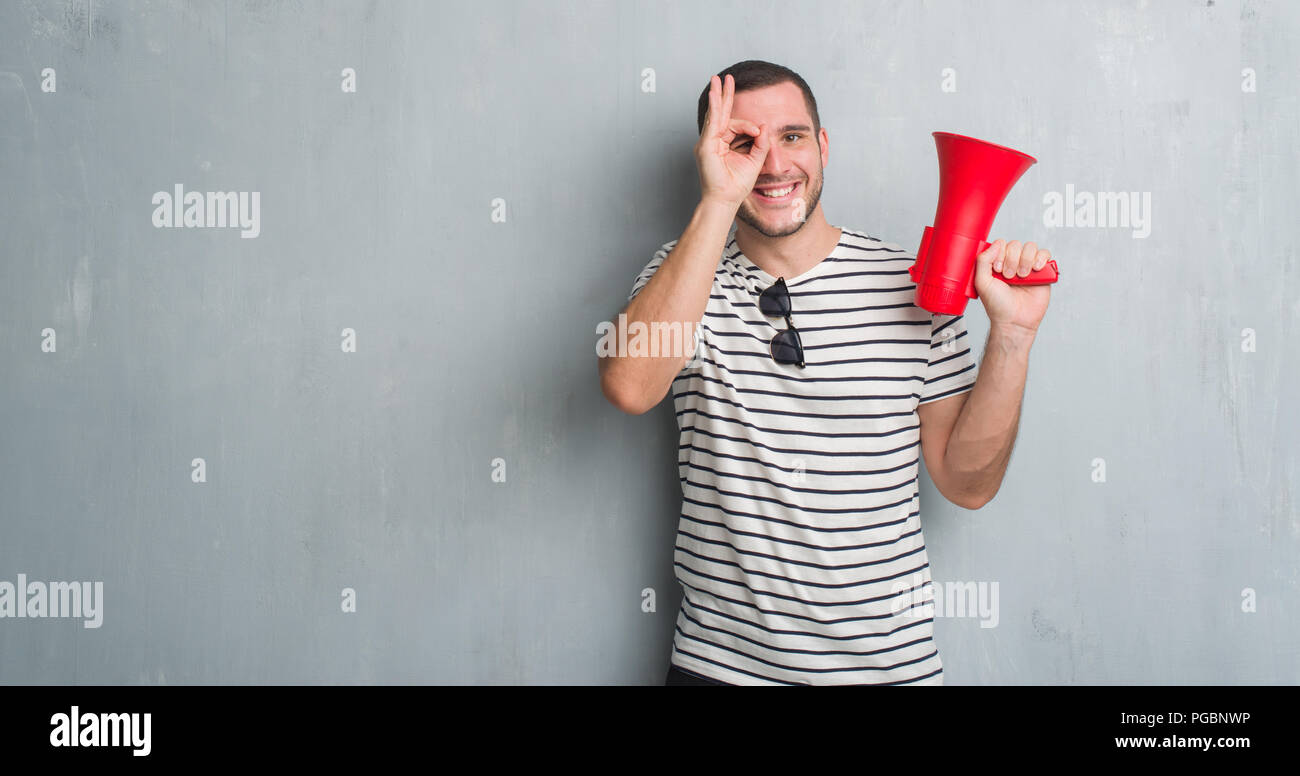 Young caucasian man over grey grunge wall shouting trough megaphone ...