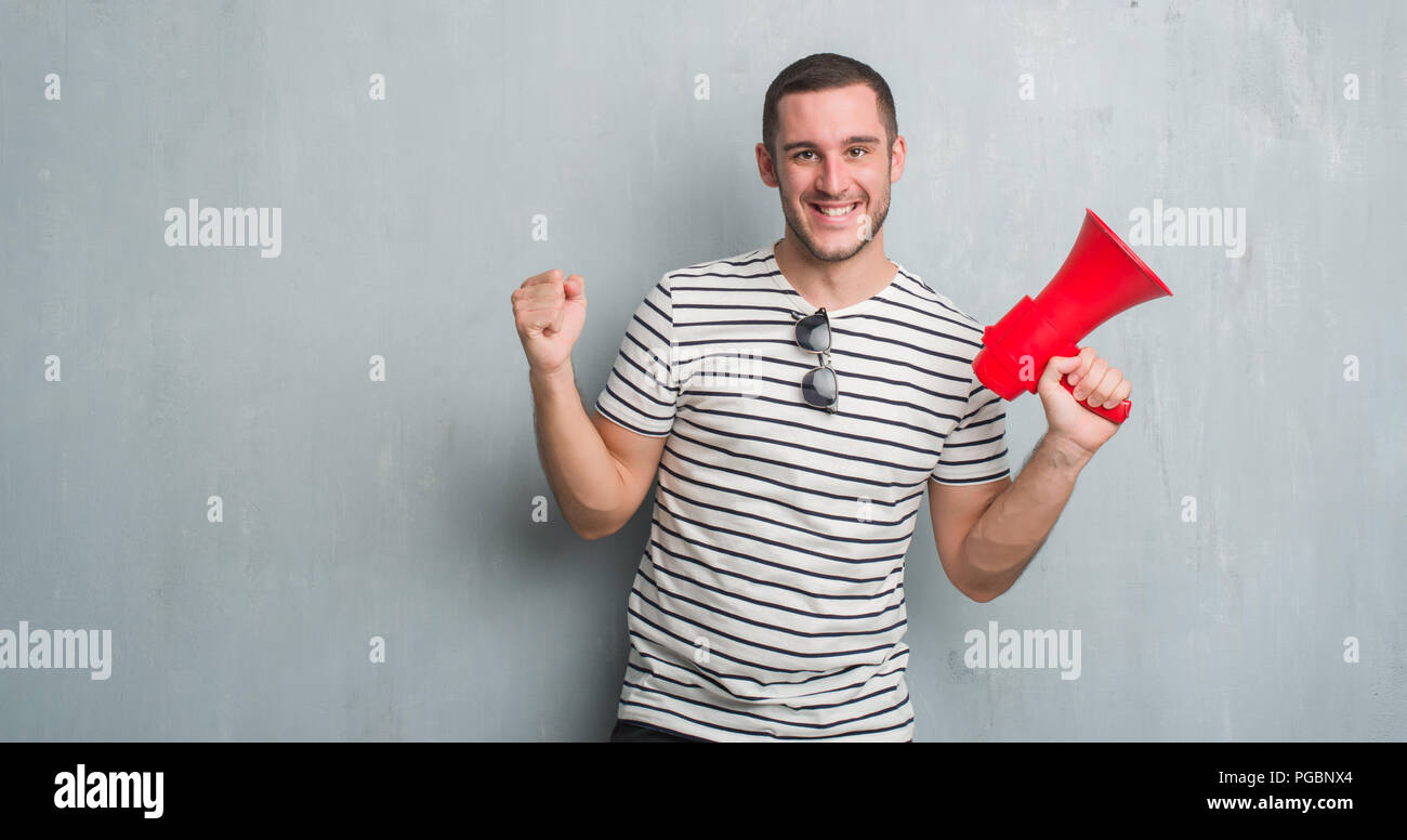 Young caucasian man over grey grunge wall shouting trough megaphone ...