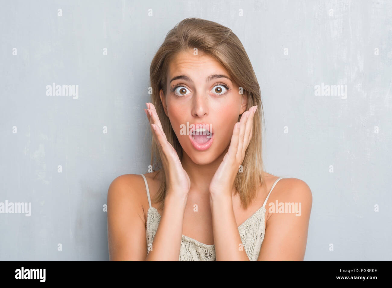 Beautiful young woman standing over grunge grey wall scared in shock ...