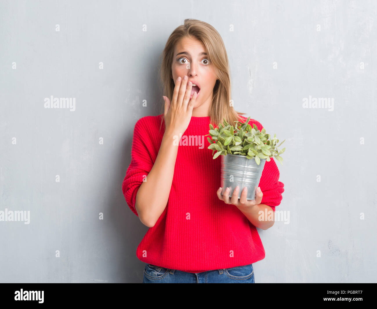 Beautiful young woman over grunge grey wall holding plant pot scared in ...