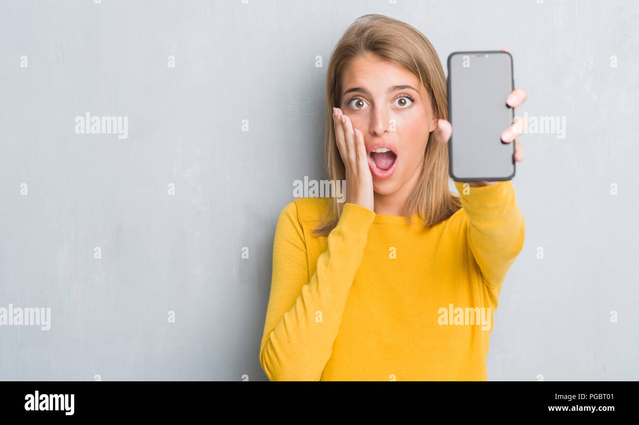 Beautiful young woman over grunge grey wall showing smartphone screen ...
