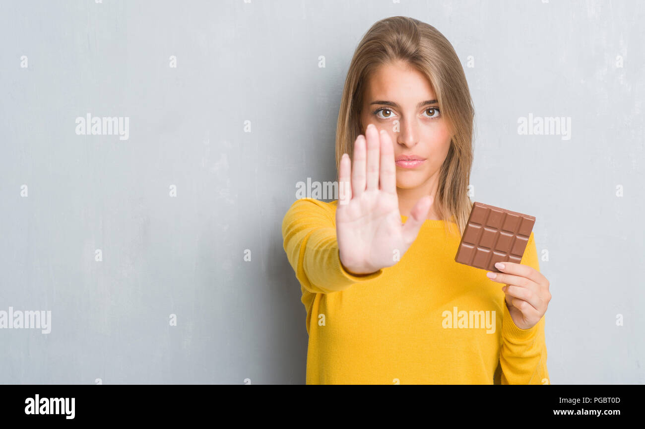 Beautiful young woman over grunge grey wall eating chocolate bar with ...