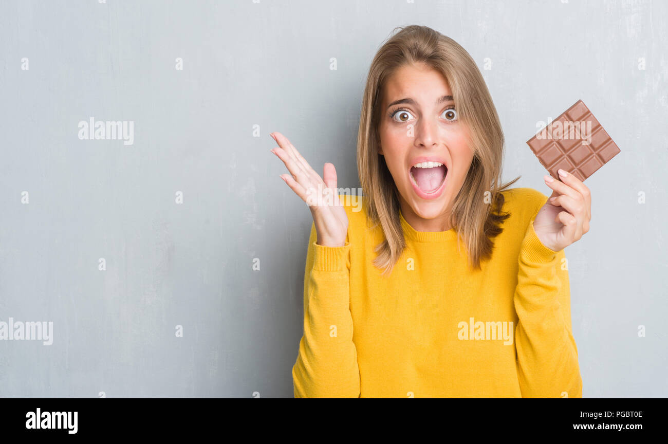 Beautiful young woman over grunge grey wall eating chocolate bar very ...