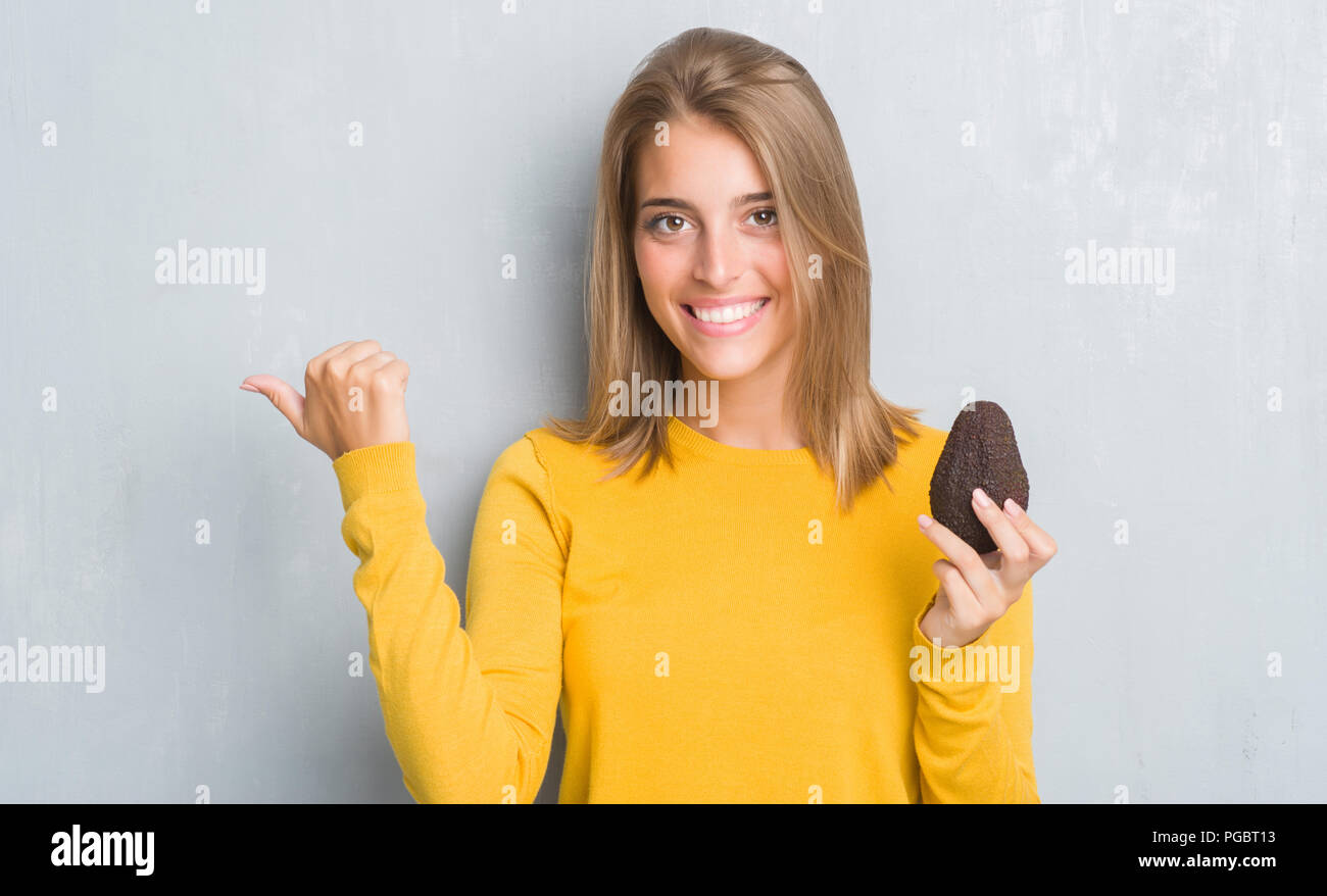 Beautiful young woman over grunge grey wall eating avocado pointing and ...