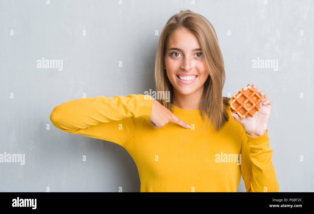 Beautiful young woman over grunge grey wall eating waffle with surprise ...