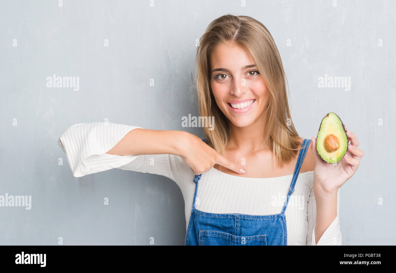 Beautiful young woman over grunge grey wall eating avocado with ...