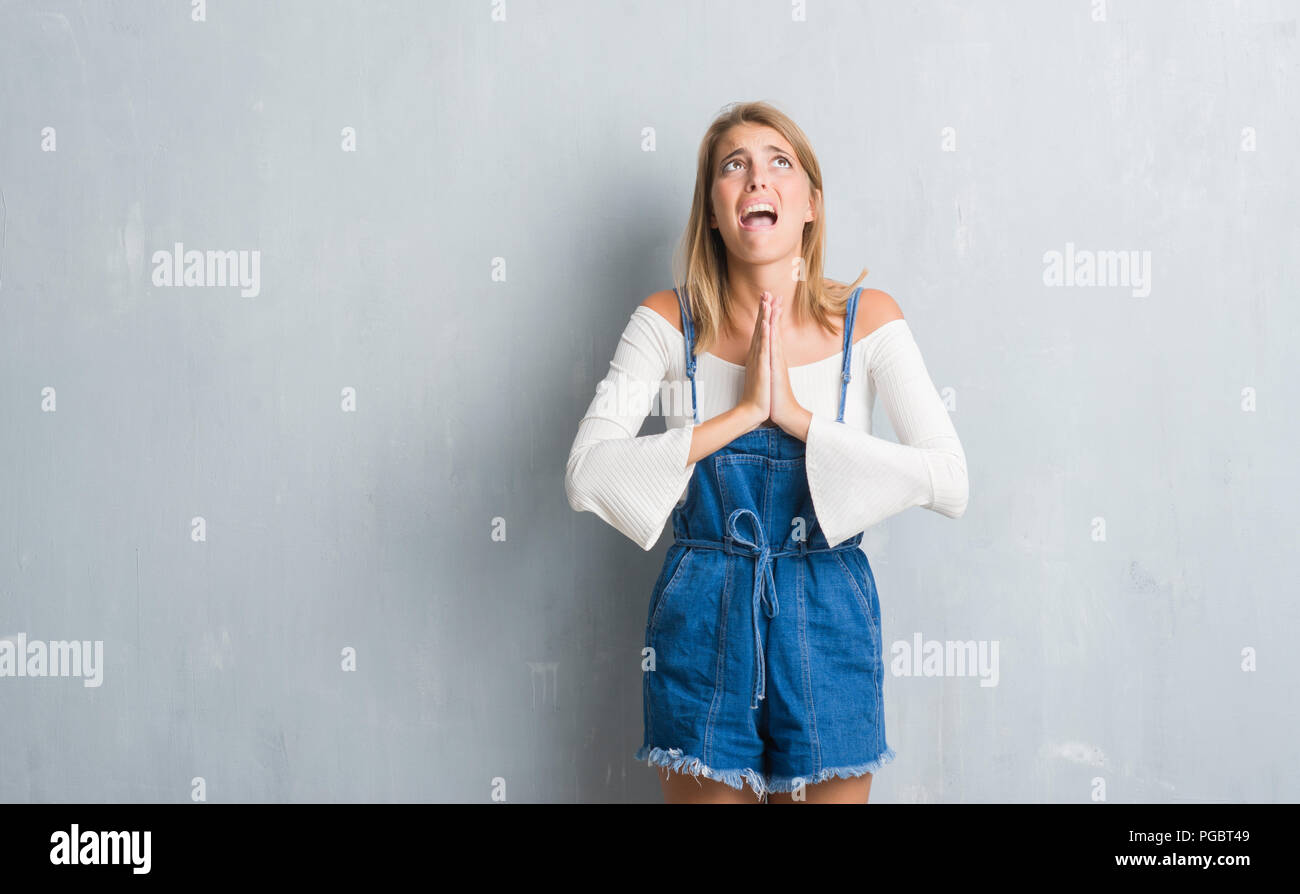 Beautiful young woman standing over grunge grey wall begging and ...