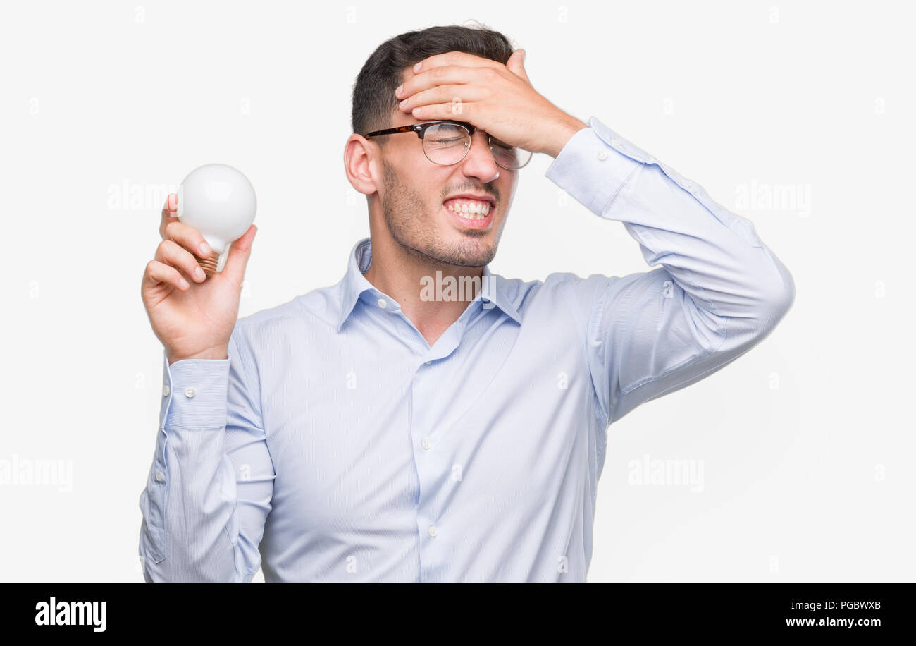 Handsome young man holding lightbulb as idea stressed with hand on head ...