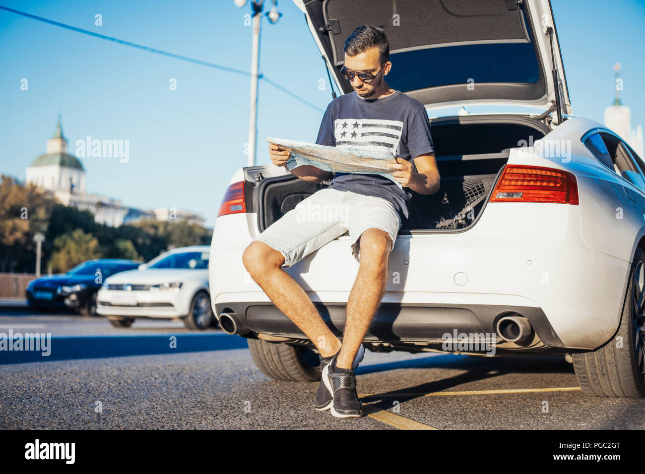 Distant plan of young man looking at map sitting in the truck of car ...