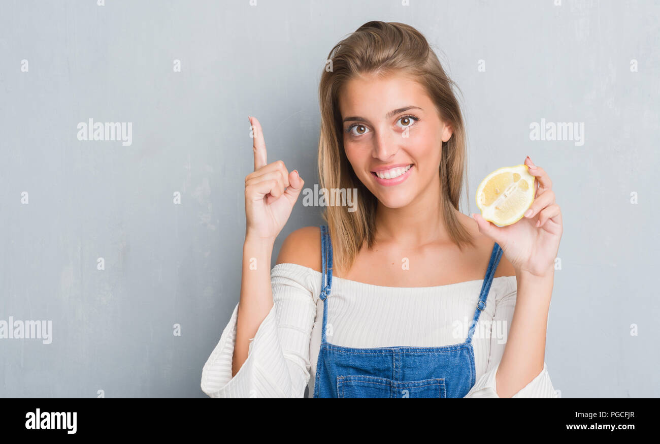 Beautiful young woman over grunge grey wall holding a lemon surprised ...