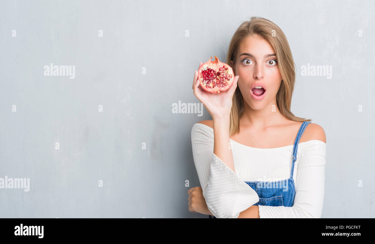 Beautiful young woman over grunge grey wall holding pomegranate scared ...