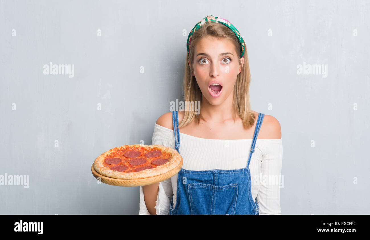 Beautiful young woman over grunge grey wall eating pepperoni pizza ...