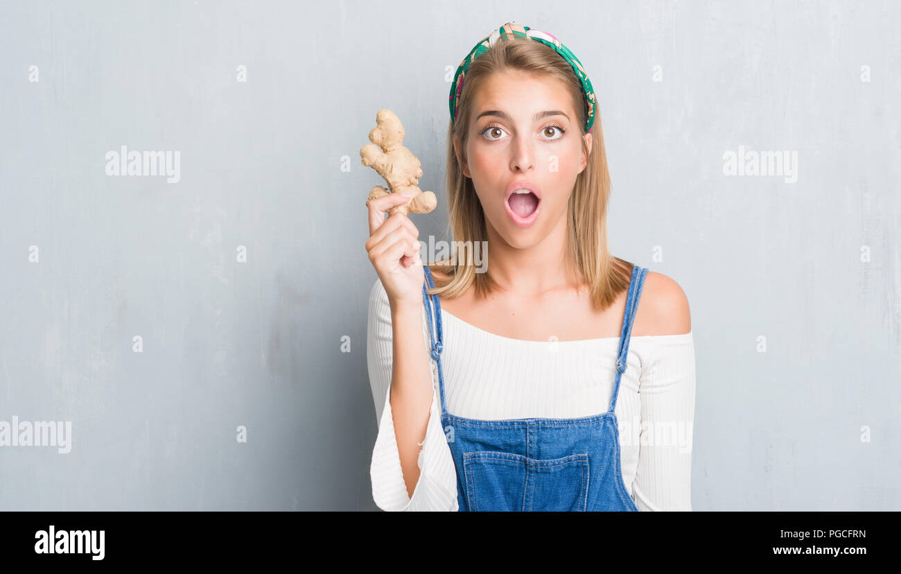 Beautiful young woman over grunge grey wall holding fresh ginger scared ...