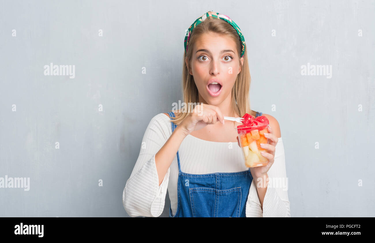 Beautiful young woman over grunge grey wall eating fruits scared in ...