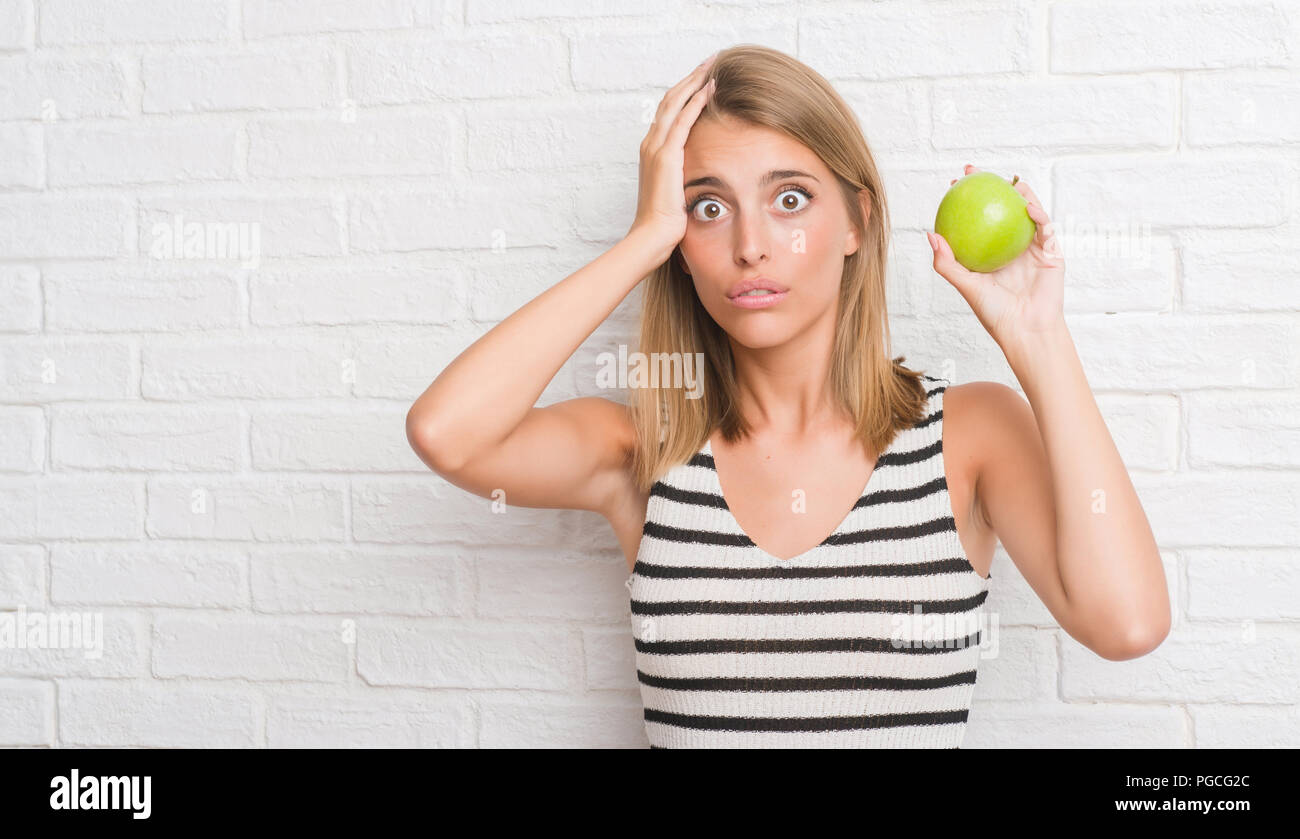 Beautiful young woman over white brick wall eating green apple stressed ...