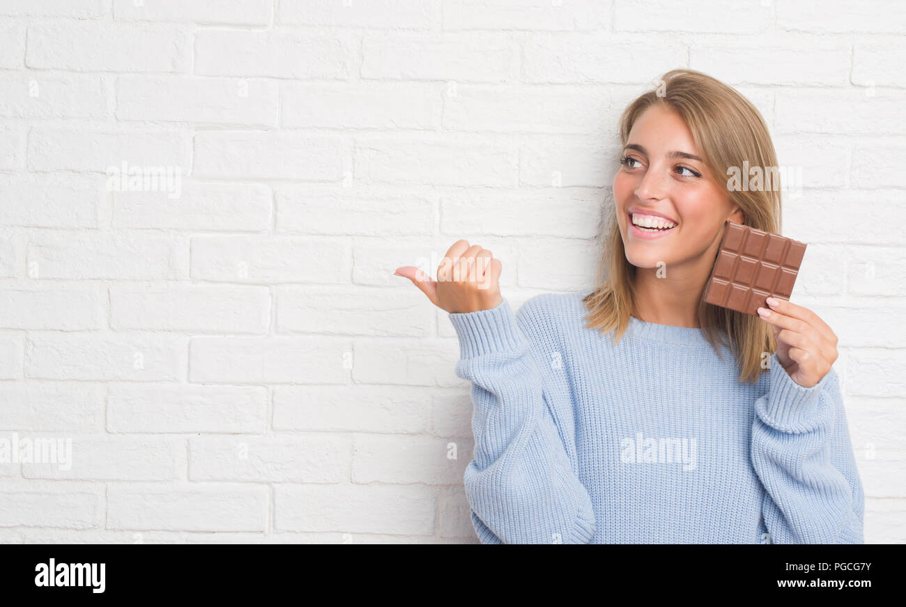 Beautiful young woman over white brick wall eating chocolate bar ...