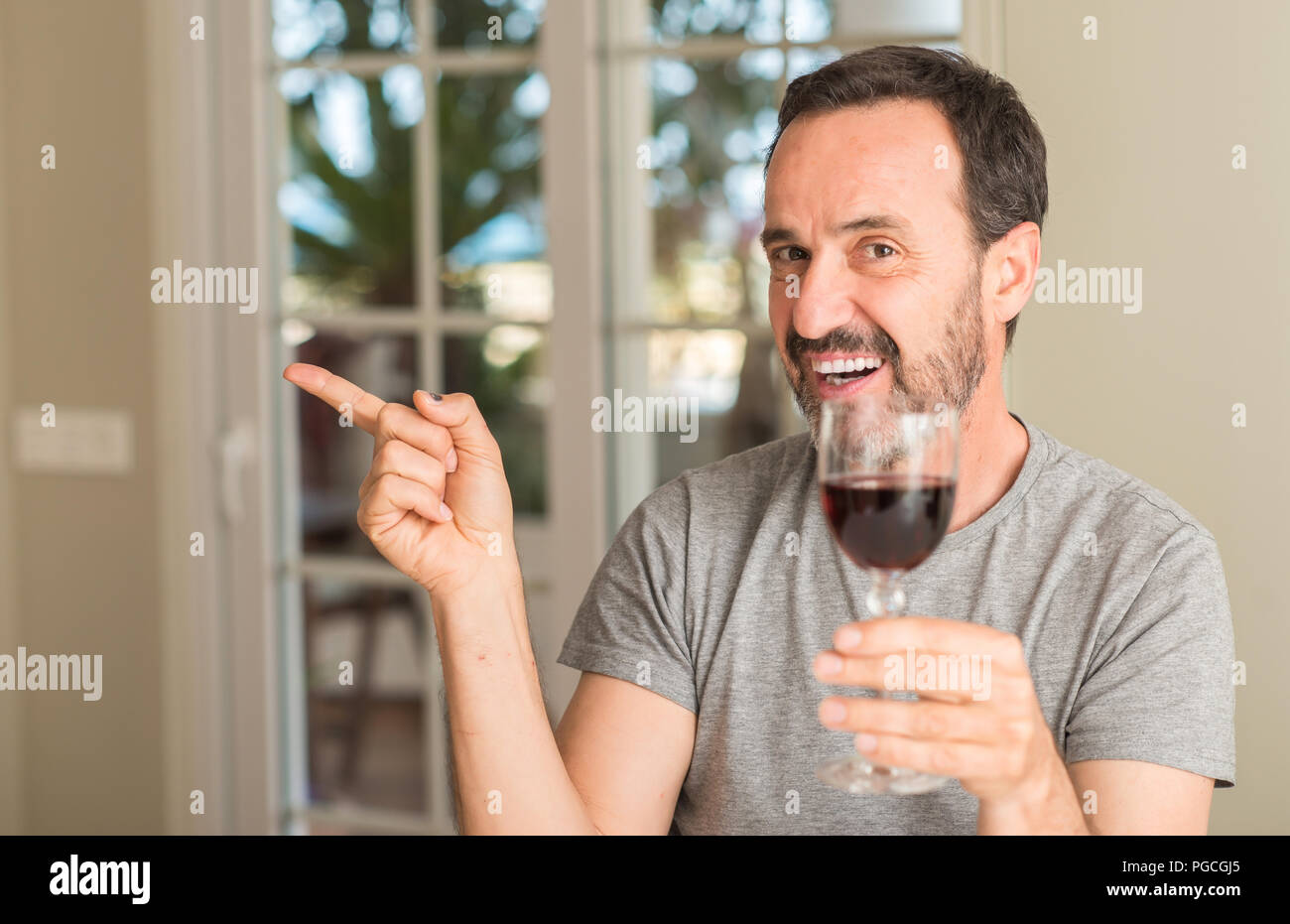 Middle age man drinking a glass of wine very happy pointing with hand ...