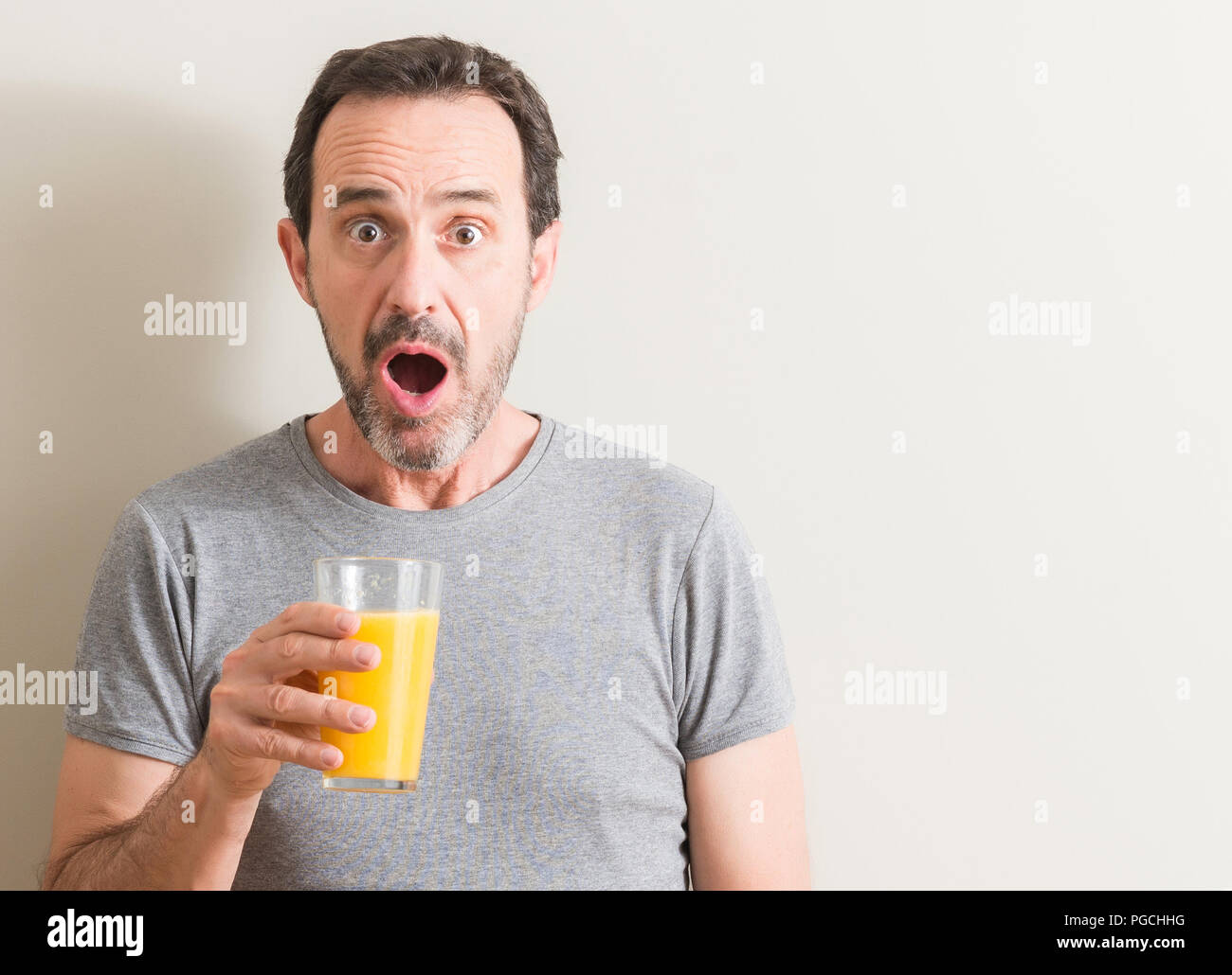 Senior man drinking orange juice in a glass scared in shock with a ...