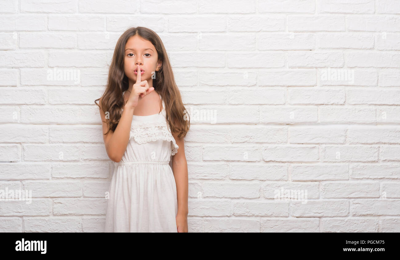 Young hispanic kid over white brick wall asking to be quiet with finger ...