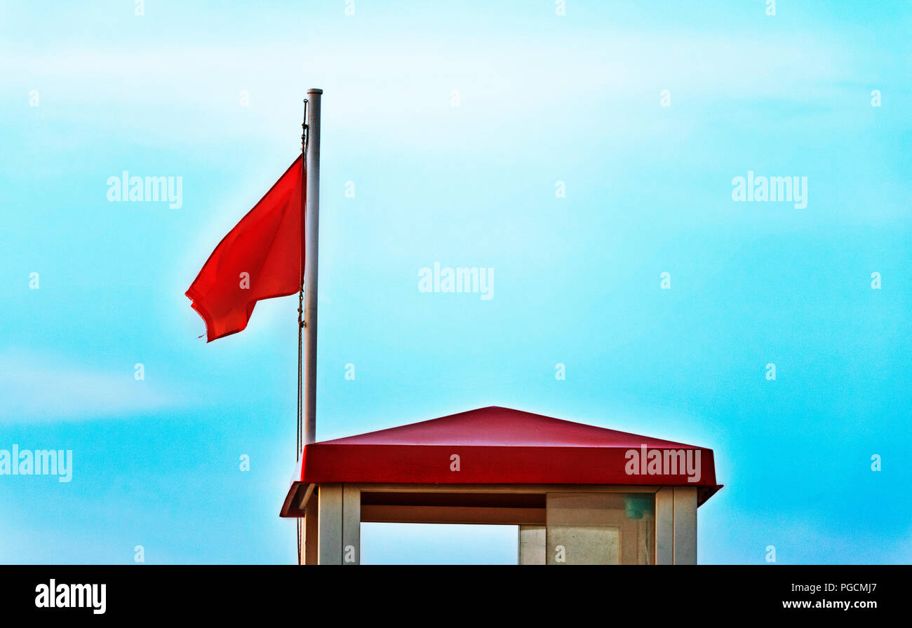 A bright red warning flag near a lifeguard tower , blue and cloudy sky ...
