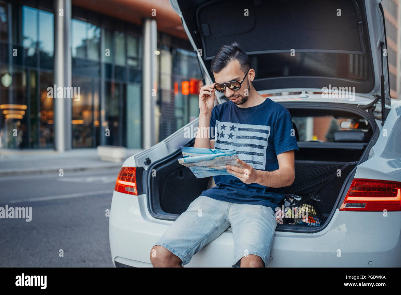 Distant plan of young man looking at map sitting in the truck of car ...