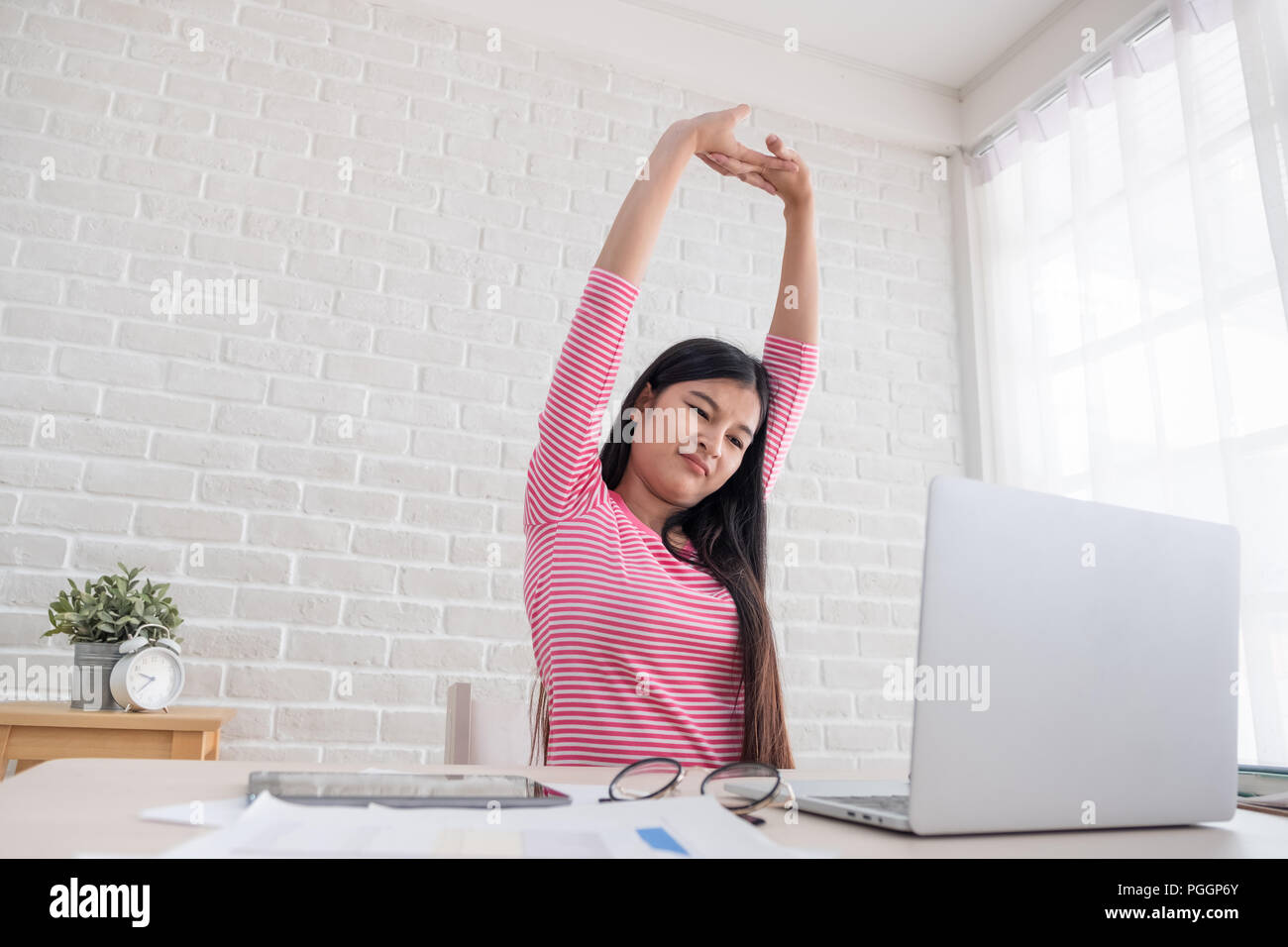 Asian woman stretching arm up when working on laptop at white brick ...