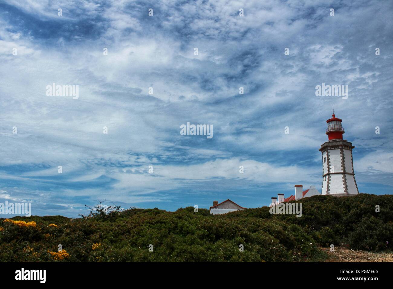 Lighthouse on top of cliff and surrounded by vegetation under cloudy sky at Cape Espichel ...