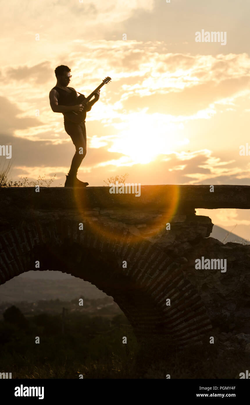 Man performs on electric guitar on old roman aqueduct during sunset ...