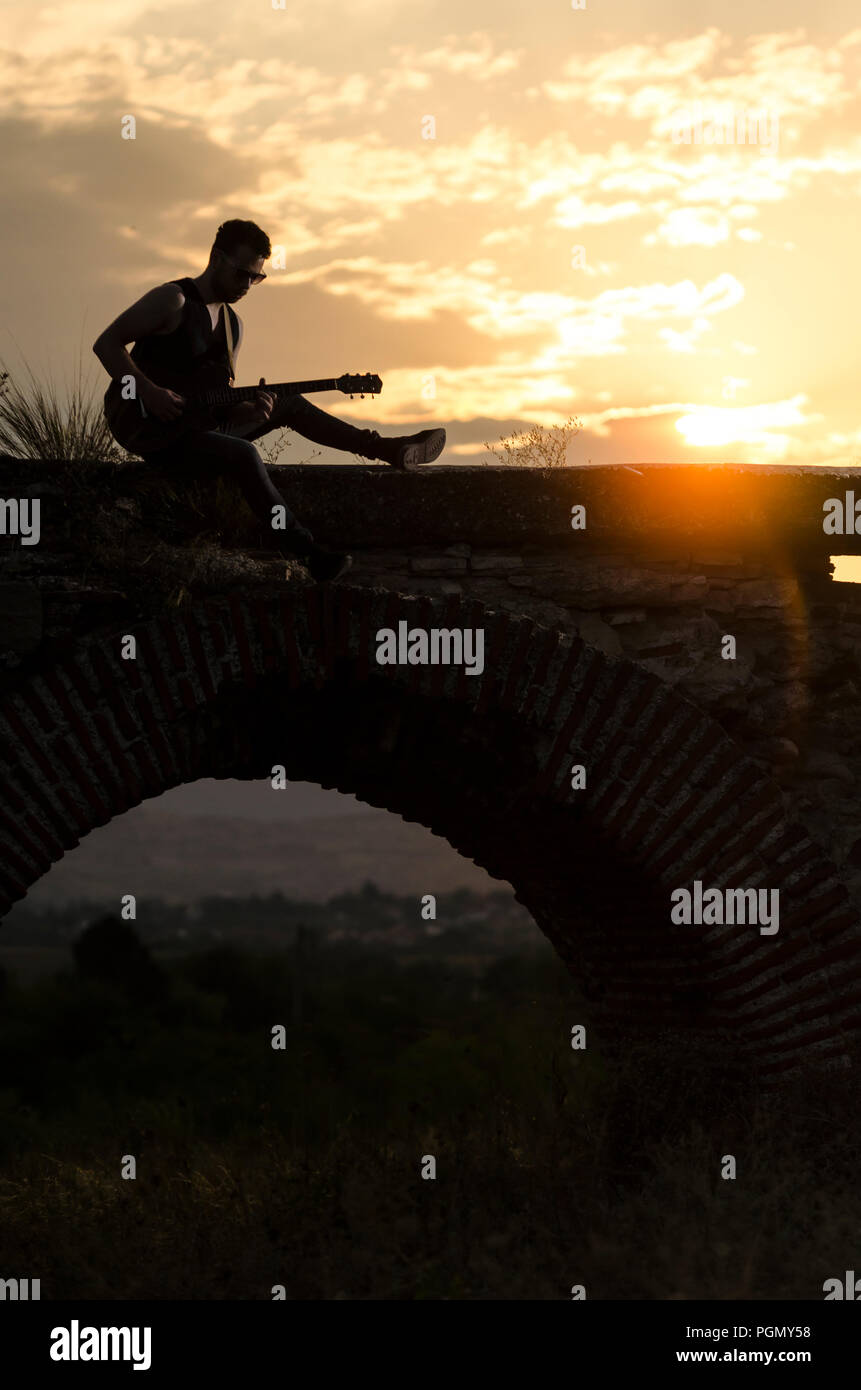 Man performs on electric guitar on old roman aqueduct during sunset ...