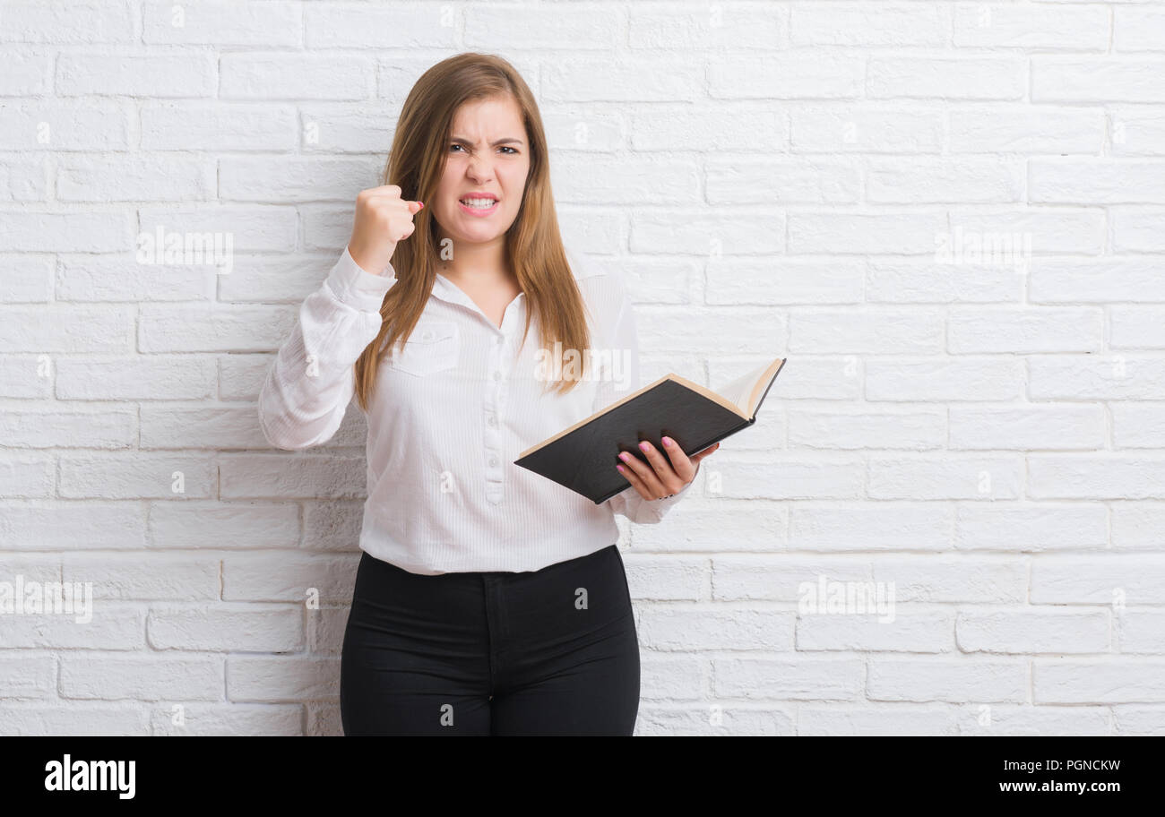 Young adult woman standing over white brick wall reading a book annoyed ...