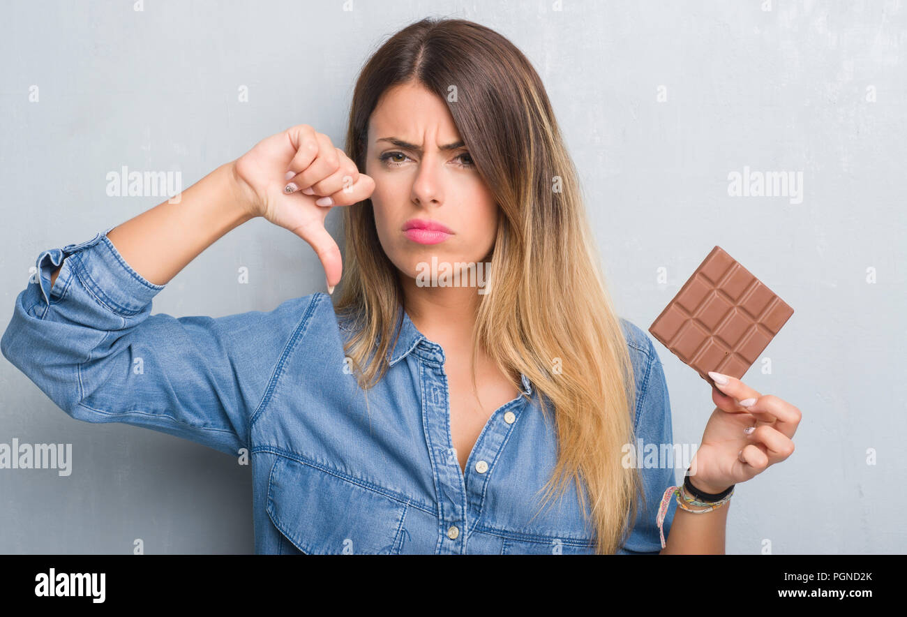 Young adult woman over grey grunge wall eating chocolate bar with angry ...