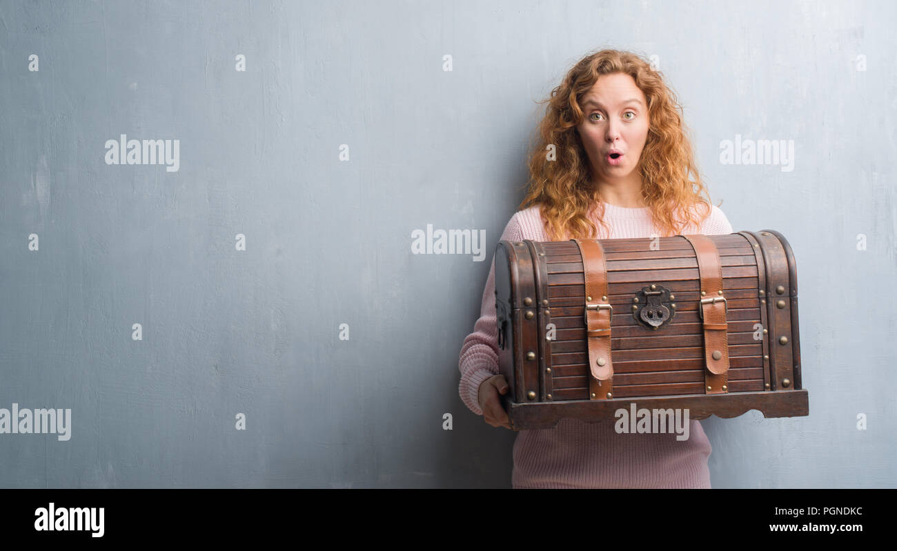 Young redhead woman holding vintage chest scared in shock with a ...