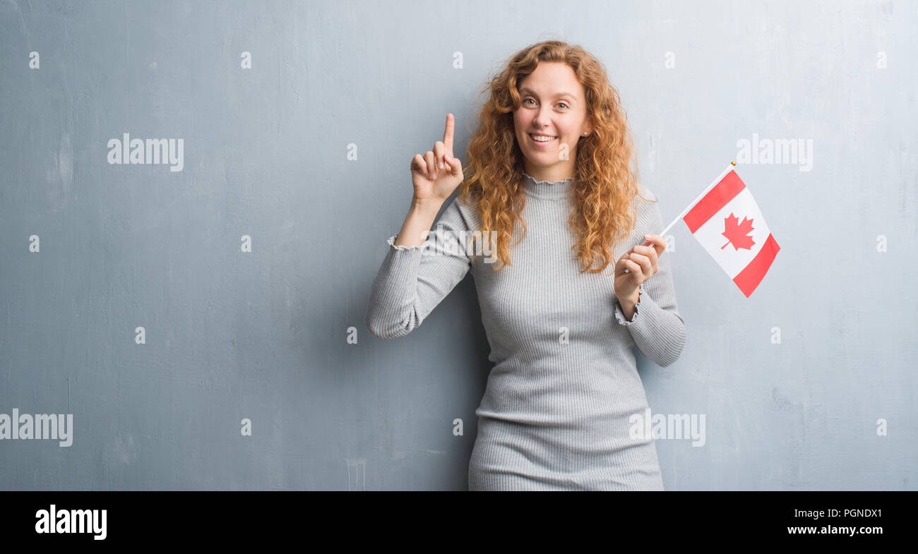 Young redhead woman over grey grunge wall holding flag of Canada ...