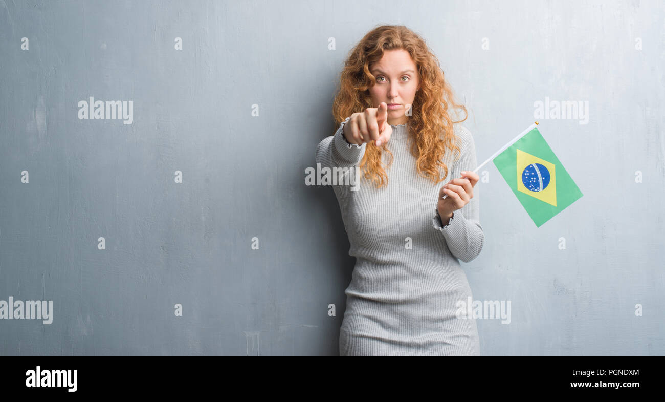 Young redhead woman over grey grunge wall holding flag of Brazil ...