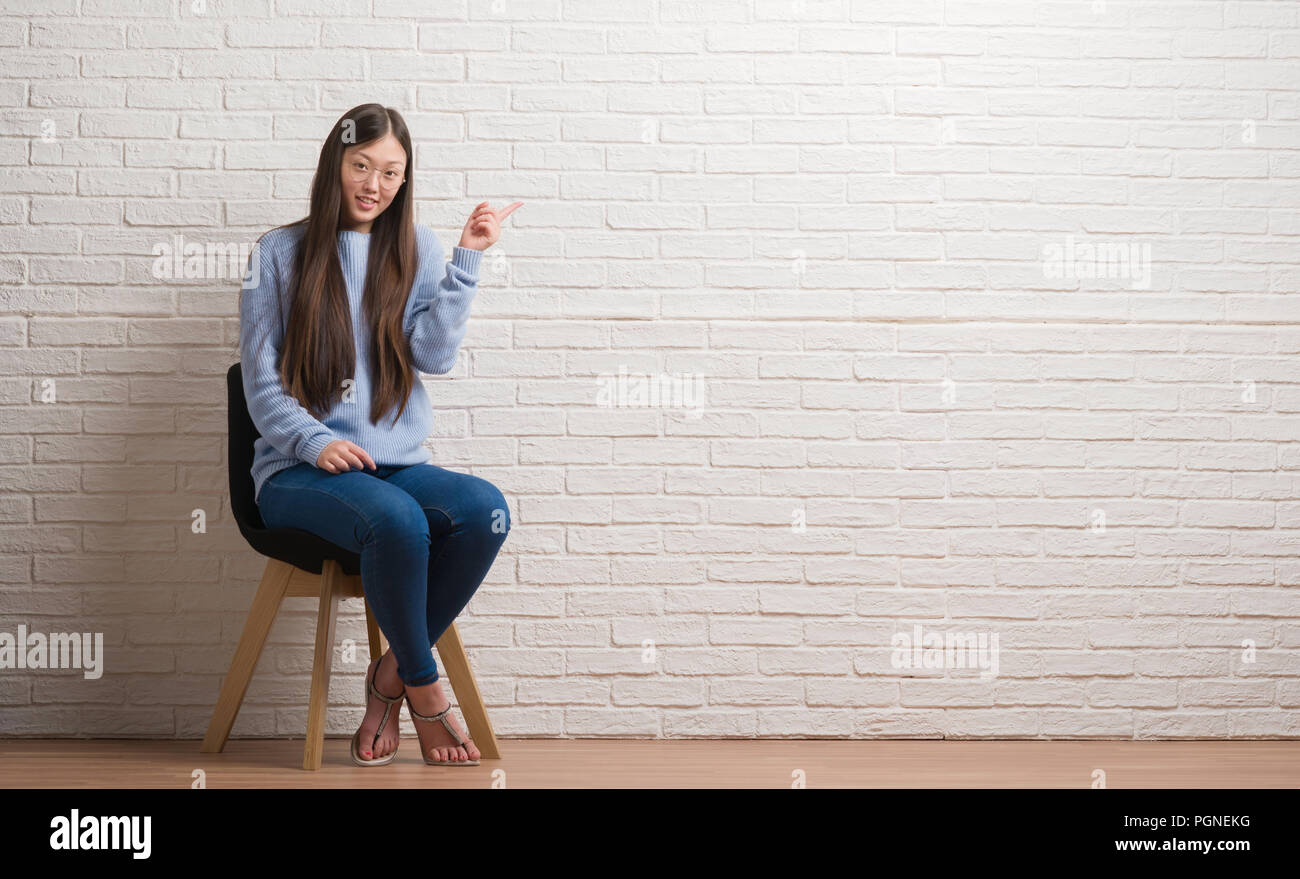 Young Chinese woman sitting on chair over brick wall very happy ...