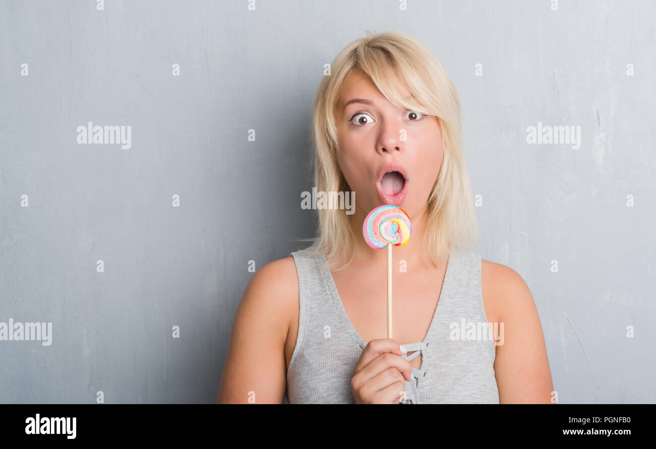 Caucasian adult woman over grey grunge wall eating candy lollipop ...
