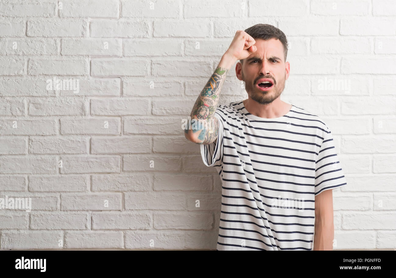 Young adult man standing over white brick wall annoyed and frustrated ...