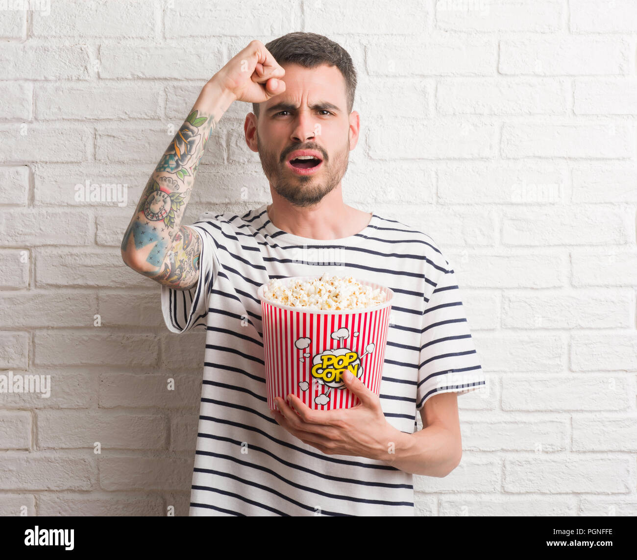 Young adult man eating popcorn standing over white brick wall annoyed ...