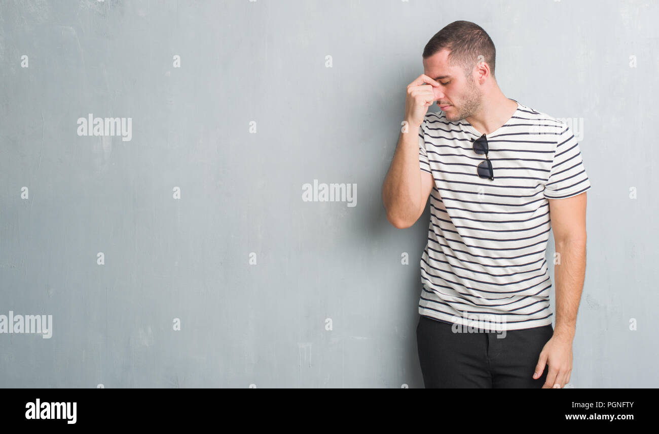 Young caucasian man over grey grunge wall tired rubbing nose and eyes ...