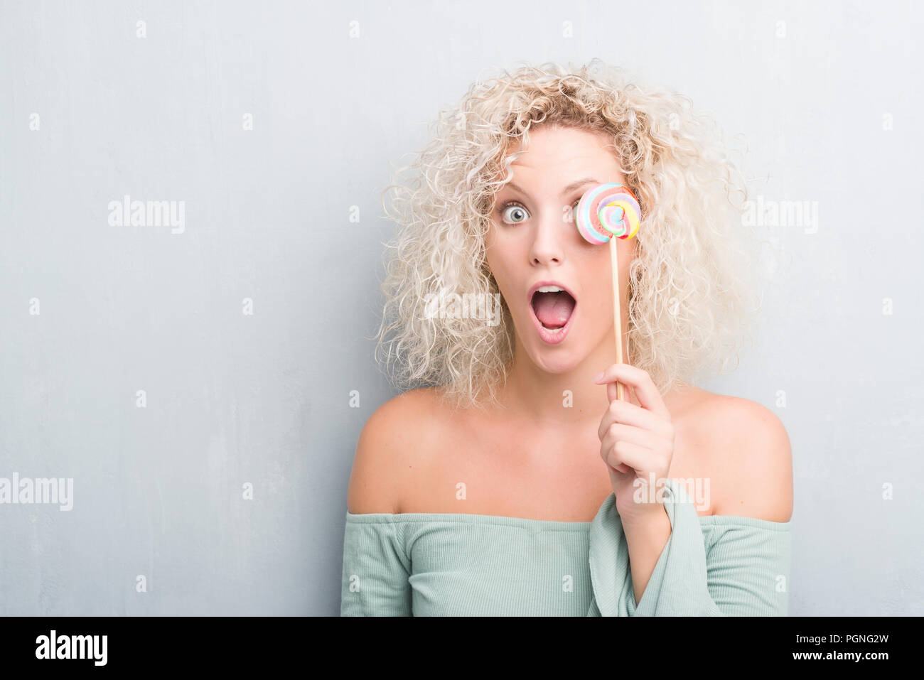 Young blonde woman over grunge grey wall eating candy lollipop scared ...
